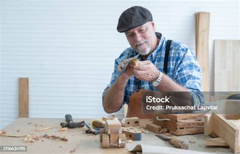 백인 시니어 늙은 흰 수염 남자 목수 앞치마와 모자 작업장에서 일하고 사포 연마 수제 나무 장난감 도구 기계 및 작은 나무 모델 또는 수공예품을 사용하여 테이블에 있습니다