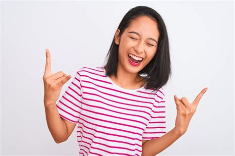 스트라이프 티셔츠를 입은 젊은 중국 여성이 고립된 흰색 배경 위에 서서 미친 표정으로 손을 들고 바위 기호를 하고 있습니다 음악