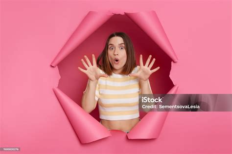 줄무늬 상의를 입고 찢어진 분홍색 종이 구멍을 통해 바라보는 재미있는 갈색 머리 여자는 누군가를 놀라게 하려고 야유를 하며 발톱을 만들며 손을 들었다 20 24세에 대한