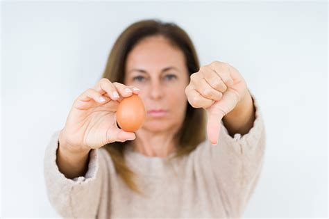 화난 얼굴로 고립 된 배경 위에 신선한 달걀을 들고 중간 나이 여자 아래로 엄지 손가락으로 혐오를 보여주는 부정적인 기호 거부