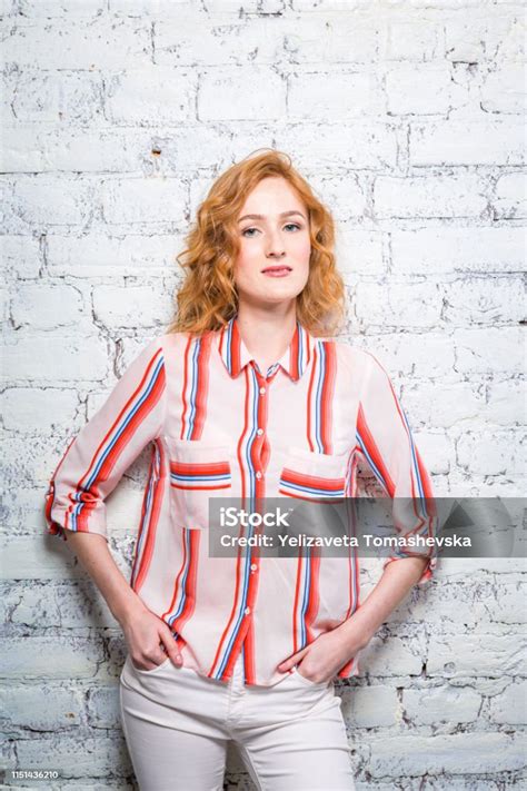 붉은 곱슬 머리와 주 근 깨와 아름 다운 젊은 여자 학생 회색 색상의 벽돌 벽에 기대어 있다 빨간 줄무늬 셔츠와 흰 바지를 입은 관능에 대한 스톡 사진 및 기타 이미지