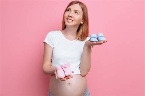 분홍색 배경에 스튜디오에서 포즈를 취하는 파란색과 분홍색 아기 부츠를 들고있는 아름다운 펜시브 임산부 귀여운에 대한 스톡 사진