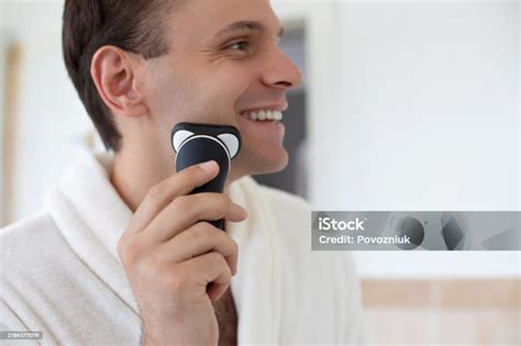 흰색 목욕 가운을 입고 웃는 남자의 클로즈업 초상화는 전기 면도기를 사용하여 턱선에 초점을 맞춰 면도합니다 그는 밝고 현대적인 욕실에서 편안한 아침 그루밍 루틴을 즐기고