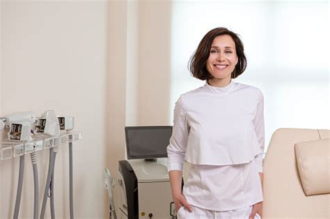 비즈니스 초상화 자신감 넘치는 여성 의사가 레이저 회춘 뷰티 트리트먼트 살롱에서 인턴스 펄스 라이트 장치에 맞서 앞쪽에 접힌 손으로 의사의 스모크에서 포즈를 취합니다 갈색