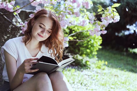 흰색 T 셔츠와 회색 치마에 젊은 아름 다운 빨간 머리 소녀 화창한 봄 꽃 벚꽃 나무 정원에서 그녀의 Blogg 채널에 대 한 책을 읽습니다 취미 블로그 블로거 아날로그