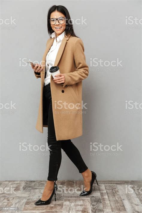 회색 배경에 고립 된 휴대 전화와 테이크 아웃 커피의 종이 컵을 들고 발 뒤꿈치에 걷는 아우터웨어를 입은 아름다운 여성의 전체