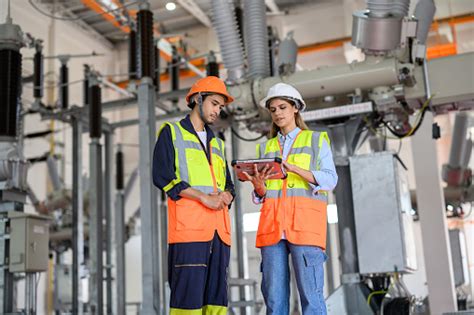 백인 전기 여성 엔지니어와 남성 아시아 기술자는 디지털 태블릿 체크리스트를 사용하여 개인 안전 장비를 사용하여 고전압 변압기 변전소의 고전압 회로 차단기 스위치의 작동 또는