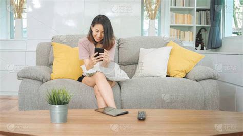 年轻的亚洲女人在家里客厅里躺在沙发上时，用智能手机检查社交媒体，笑得很开心。生活方式，拉丁裔和他的恐