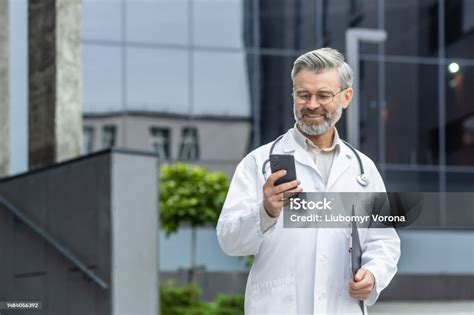 흰 코트를 입은 남성 선임 의사가 청진기와 손에 팩을 들고 진료소 밖에 서서 전화를 사용하고 메시지를 쓰고 화상 통화로 이야기하고 미소를 짓습니다 의사에 대한 스톡 사진 및
