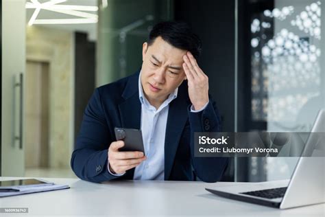 불만족스러운 아시아 사업가는 나쁜 소식을 듣고 노트북으로 현대적인 사무실 안에 앉아 스마트폰으로 읽는 남자 전화 화가 난 실망한