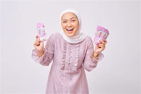 히잡과 보라색 드레스를 입고 흥분한 아름다운 아시아 무슬림 여성이 흰색 스튜디오 배경에 고립된 카메라를 바라보며 루피아 지폐에 현금을 들고 있다 Eid Ul Fitr에 대한