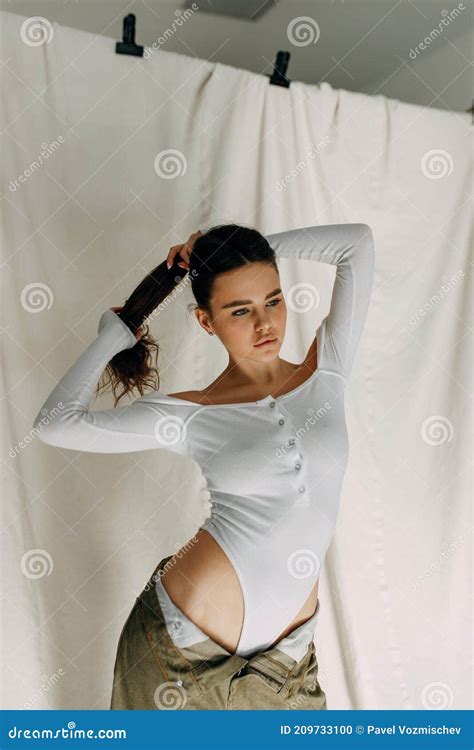 Portrait Of A Young Skinny Brunette Posing In A White Bodysuit Sitting On A White Floor In A