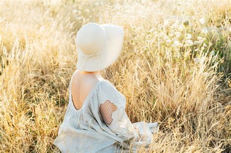 가벼운 바람이 잘 통하는 드레스를 입고 등을 열고 밀짚모자를 쓴 부드러운 뒷모습 여성이 해질녘 높은 풀밭에서 순간을 즐기고 있다 자연의 여름 저녁 차분하고 조화 몸에 주근깨