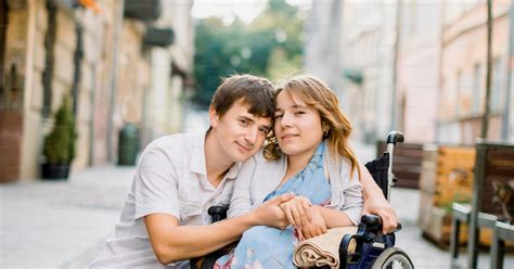 휠체어를 탄 예쁜 젊은 웃는 여자와 카메라를 보고 있고 손을 잡고 사랑에 빠진 잘생긴 남자가 오래된 도심을 걷고 있다 사진 Unsplash의 사람 이미지