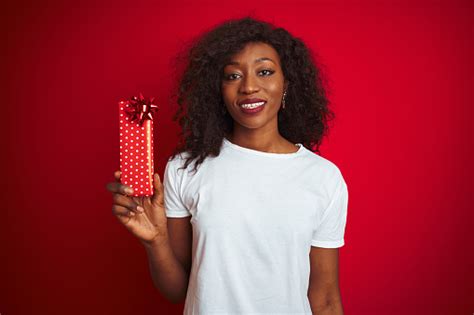 행복한 얼굴 서서 치아를 보여주는 자신감 미소와 함께 미소와 고립 된 빨간 배경 위에 서 발렌타인 선물을 들고 젊은 아프리카 계 미국인 여성 건강한 생활방식에 대한 스톡