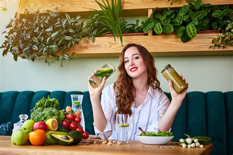 배경에 녹색 꽃과 신선한 과일과 야채와 아름다운 인테리어에 스무디와 신선한 물을 마시는 젊은 여성 건강한 식생활 개념 비건 식사