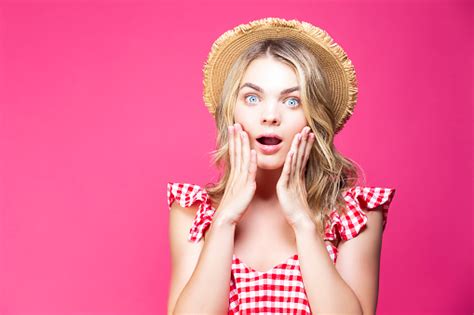 산호 티셔츠와 밀짚모자를 쓴 금발 소녀가 여름 반바지를 입고 포즈를 취하며 산호 분홍색 배경 위에 놀란 표정으로 뺨을 만지고 있다 금발 머리에 대한 스톡 사진 및 기타