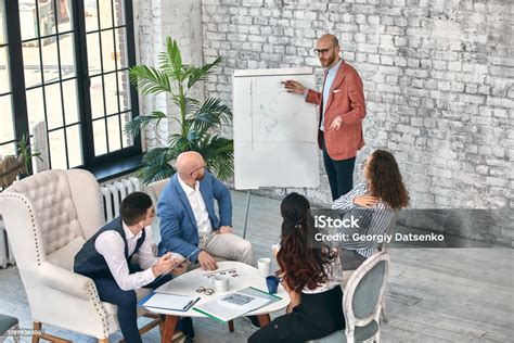 플립 차트에 글을 쓰는 자신감 있는 사업가는 회사 회의에서 회의실에서 새로운 프로젝트를 발표합니다 진지한 멘토 감사관이 이사회 와 그래프를 사용하여 비즈니스에 대해 파트너와