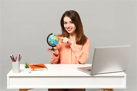 지구 세계 글로브에 인덱스 손가락을 가리키는 젊은 웃는 여자 앉아서 회색 배경에 고립 된 Pc 노트북으로 흰색 책상에서 작동 합니다 성취 비즈니스 커리어 라이프 스타일 컨셉