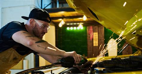 차고에서 작업하는 동안 모터의 세부 사항을 수리하기 위해 전기 그라인더를 사용하는 동안 자동차 엔진 위로 구부러진 젊은 수리공