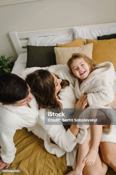 젊은 엄마 아버지는 침대에 누워 목욕 가운을 입은 귀여운 딸을 껴안고 호텔 방에서 서로를 바라본다 행복한 엄마 아빠 여자 아이가