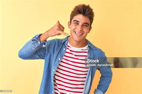 줄무늬 티셔츠와 데님 셔츠를 입은 젊은 잘생긴 남자는 고립 된 노란색 배경 위에 전화와 같은 손과 손가락으로 전화 제스처를하고 미소 개념을 전달합니다 T 셔츠에 대한 스톡