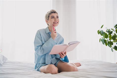 잠옷을 입고 공책을 들고 나무 침대와 미니멀한 인테리어가 있는 밝은 침실에서 사려 깊게 미소 짓는 젊은 여성 25 29세에 대한