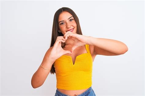 고립 된 흰색 배경 위에 서있는 노란색 캐주얼 티셔츠를 입은 어린 아름다운 소녀는 손으로 심장 기호와 모양을 보여주는 사랑에 미소지었습니다 로맨틱 한 개념 T 셔츠에 대한