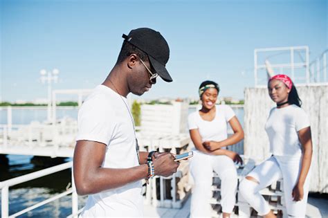 세련 된 아프리카계 미국인 소년 흰색 옷에 착용 안경 및 모자 두 여자에 대 한 손에 휴대 전화 젊은 흑인 들의 스트리트 패션