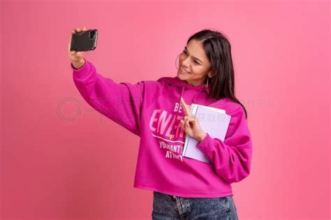 连帽衫高清图片 穿着粉色连帽衫的西班牙裔漂亮女人微笑着拿着笔记本，用智能手机摆出粉色孤立的姿势，在相机上自拍 产品实拍免费下载格式