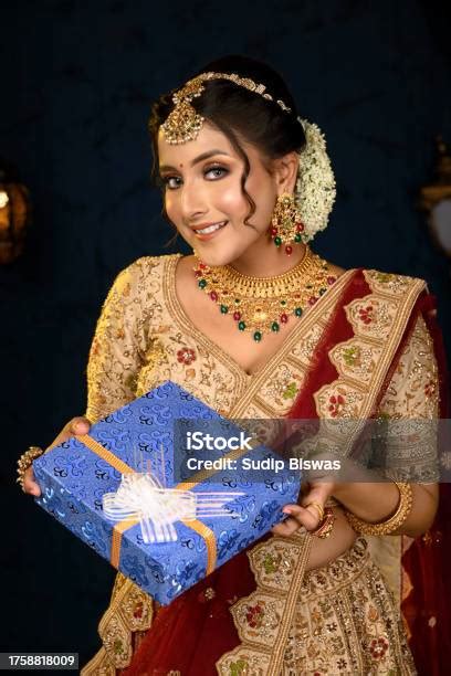 예쁜 젊은 인도 여성의 초상화는 전통적인 Lehenga 금 보석 및 팔찌를 입고 장식 배경에 손에 선물 상자를 들고 있습니다 인도 문화 행사 종교 및 패션 25 29세에
