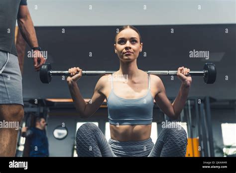 Fit Girl Doing Squats With Weights In Gym And Male Trainer Watching Her