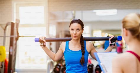 파란색 싱글릿을 입은 여자는 개인 트레이너와 함께 체육관에서 바에서 운동하고 있으며 클립보드의 운동 계획에 글을 쓰고 있다