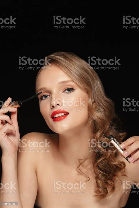 물결 모양의 머리와 빨간 입술을 들고 있는 젊은 아름다운 여성의 초상화가 검은 색 배경을 통해 카메라를 꿈꾸며 바라보며 마스카라를 들고 있습니다 16 17 살에 대한 스톡