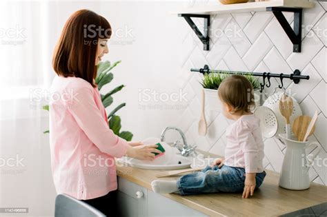 젊은 행복 한 어머니와 그녀의 귀여운 곱슬 유아 딸 흰색 부엌에서 함께 재미 어머니는 접시를 씻는 동안 아기 소녀는 싱크대
