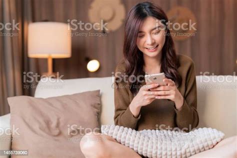 매력적인 젊은 아시아 성인 여성은 집에서 소파에서 휴식을 취하면서 휴대 전화를 사용하고 아시아 여성은 스마트 폰을 사용하여 인터넷 소셜 미디어를 서핑하거나 집에서 쾌활한