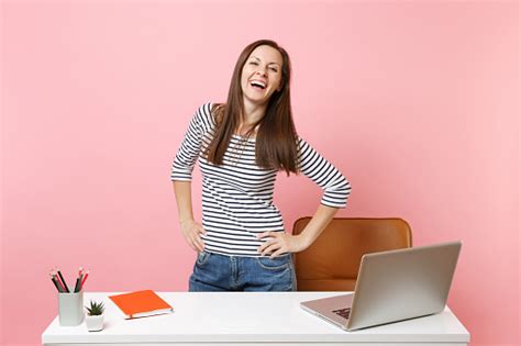 영 캐주얼 옷에서 여자 웃음 작동 파스텔 핑크 배경에 고립의 현대 Pc 노트북과 흰색 책상 근처에 서 성과 비즈니스 경력