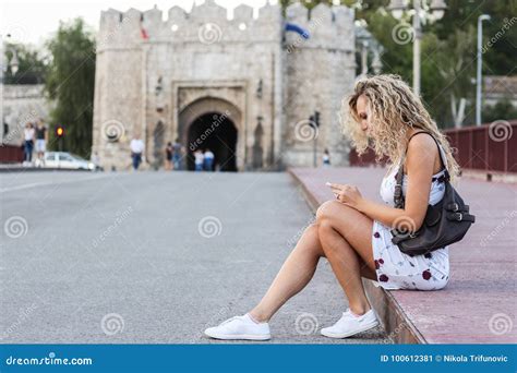 Blonde Girl In A White Dress Sitting On A Sidewalk On The Bridge Stock