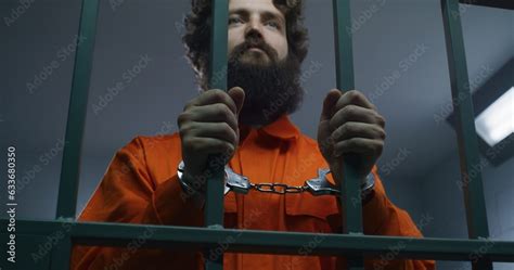 Male Prisoner In Orange Uniform Holds Metal Bars Stands In Prison Cell