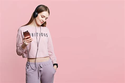 캐주얼 옷에 사랑 스러운 슬림 젊은 여성 Hearphones에 좋아하는 오디오 트랙을 즐깁니다 그리고 현대 휴대 전화를 사용 하 여 텍스트 복사 공간 핑크 배경 위에 고립