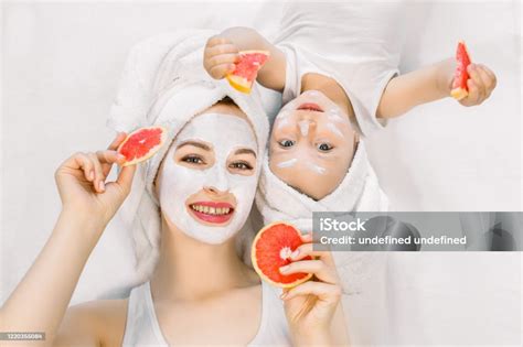 행복한 가족 어머니와 귀여운 작은 딸 흰색 티셔츠와 침대에 누워 자신의 머리에 흰색 수건 점토 마스크와 얼굴 미용 절차를하고