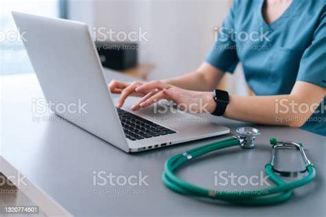 책상에 앉아 노트북 키보드에 입력 하는 의료 유니폼 작업에서 인식 할 수없는 여성 의사의 클로즈업 손 컴퓨터에 대한 스톡 사진