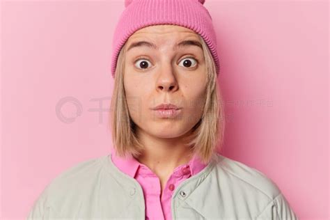 粉红色高清图片 震惊的女人的水平镜头凝视着眼睛，注意到一些可怕的东西，无法相信可怕的消息，穿着夹克帽，与粉红色背景隔离 人类反应概念 产品实拍