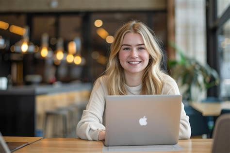 A Smiling Blonde Woman Sitting At A Table With A Laptop And Looking Into A Camera While Working