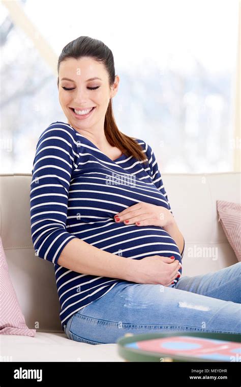 Happy pregnant woman sitting on sofa and touching her baby bump while ...
