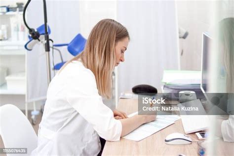 클리닉 병원에서 흰색 유니폼을 입은 산부인과 의사가 컴퓨터에서 작동합니다 배경에 의자 및 기타 의료 장비가있는 부인과 캐비닛