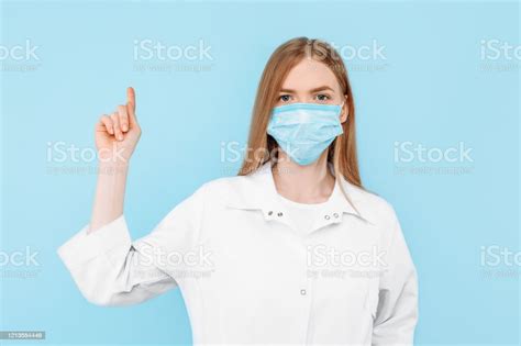 의료 용 마스크와 흰색 실험실 코트를 입은 여성 의사가 고립 된 파란색 배경을 가리켰습니다 의료의 개념 코로나바이러스 Covid