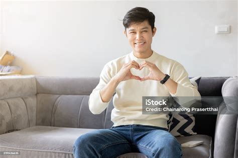 클로즈업 젊은 아시아 남자는 행복 느낌으로 미소 짓고 건강을 위해 거실의 소파에 앉아있는 동안 하트 모양 제스처를 만들고 더 나은 발렌타인 데이와 사람들 라이프 스타일 개념