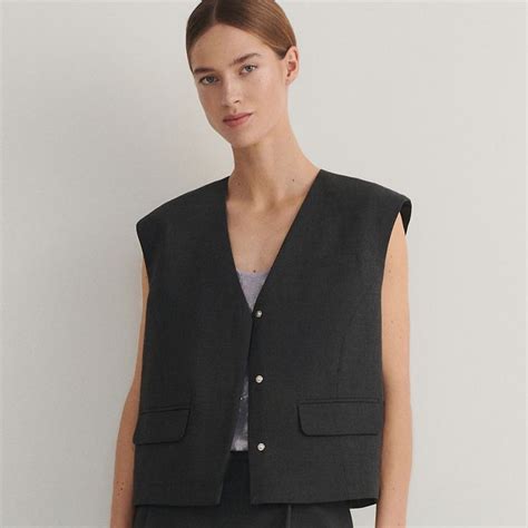 A Woman Standing In Front Of A White Wall Wearing A Black Vest And Grey Shirt