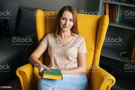 현대적인 인테리어가있는 어두운 방에서 종이 책을 들고 편안한 노란색 의자에 누워있는 예쁜 금발 여성의 높은 각도보기 가정 생활에